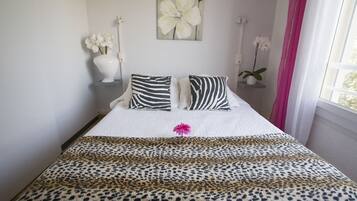 Quarto com cama, colcha de onça, duas almofadas com estampa de zebra, vaso branco com flores, pintura de uma flor e cortina rosa.