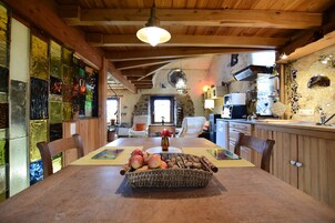 Uma mesa de jantar de madeira com uma cesta de palha contendo maçãs e nozes, um vaso com flores, e vista para uma cozinha e sala de estar com paredes de pedra e vigas de madeira.