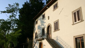 Um edifício de dois andares com uma escadaria que leva à entrada, cercado por plantas em vasos e árvores.