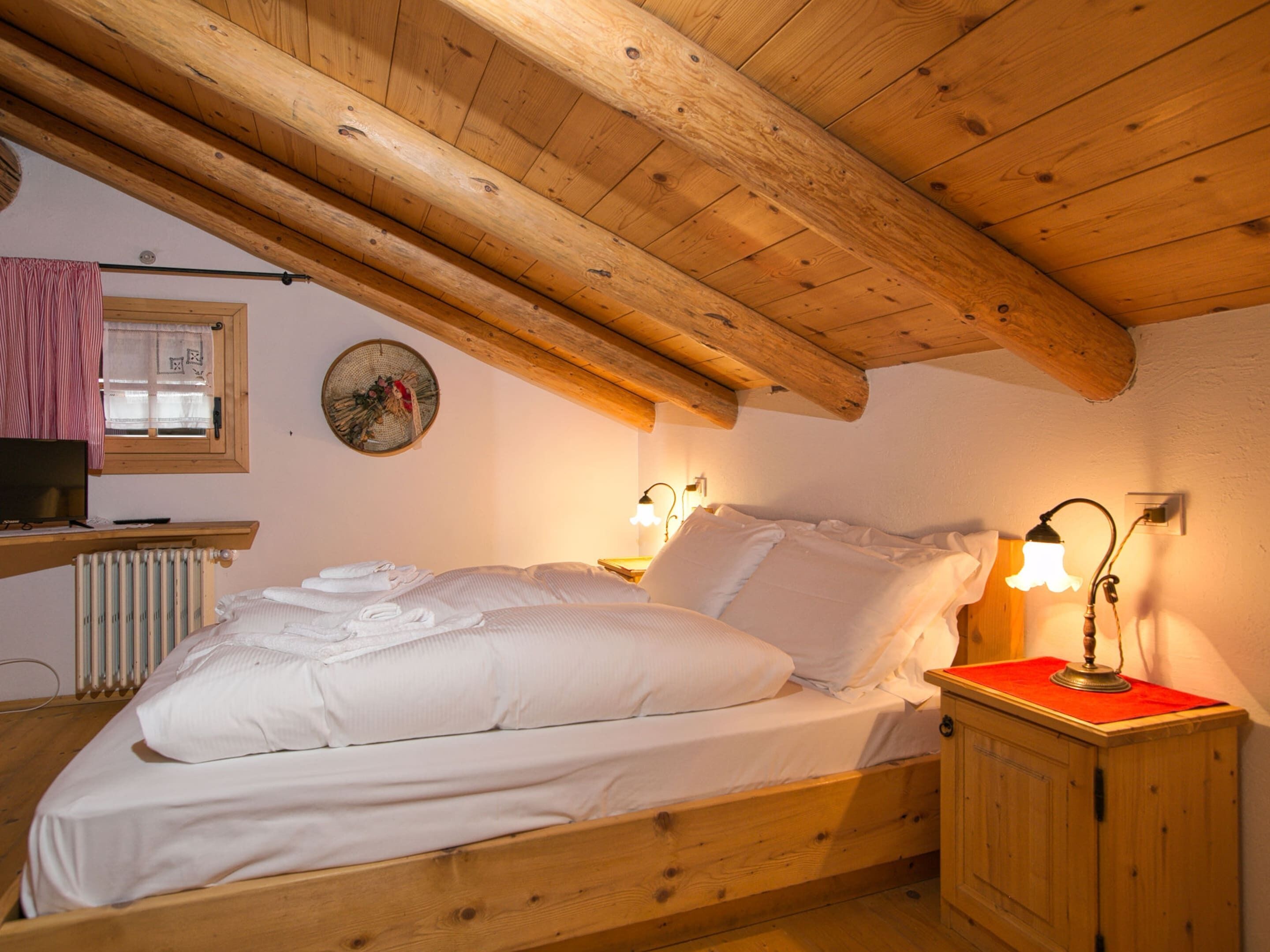 Quarto aconchegante com teto de madeira, cama com lençóis brancos, mesa de cabeceira com abajur e radiador.