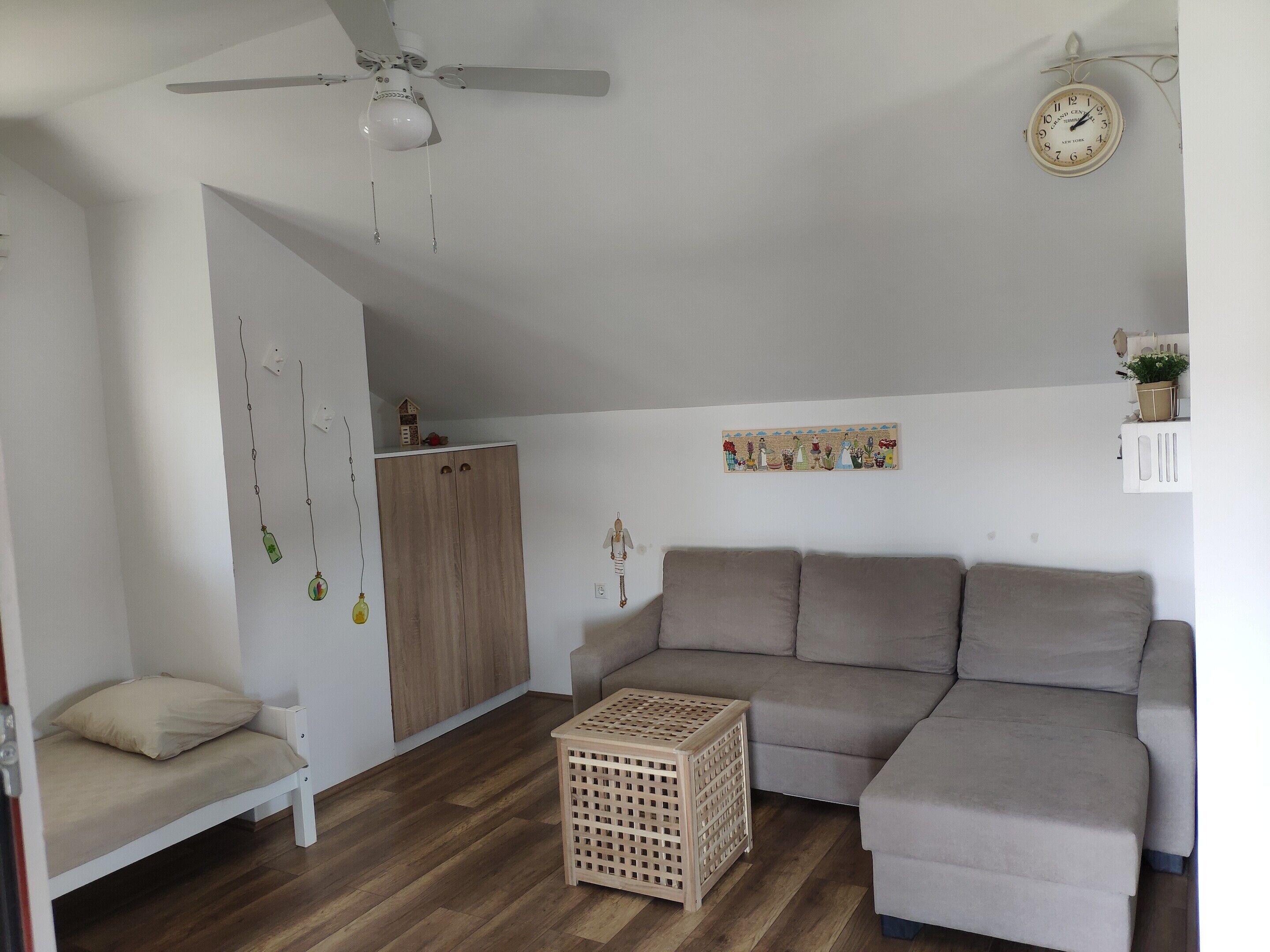 Uma sala de estar com piso de madeira, um sofá bege, uma pequena mesa de madeira, um ventilador de teto branco e um relógio de parede.