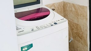 Máquina de lavar roupa compacta com porta transparente, painel de comandos e tomada na parede.
