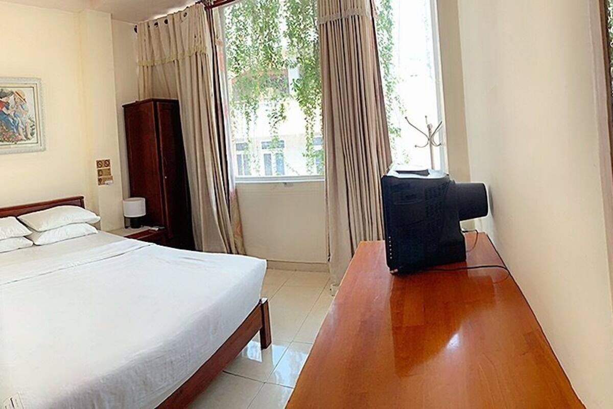Quarto de hotel com duas camas, uma mesa de madeira, uma escrivaninha, uma cadeira, uma janela com cortinas e um quadro na parede.