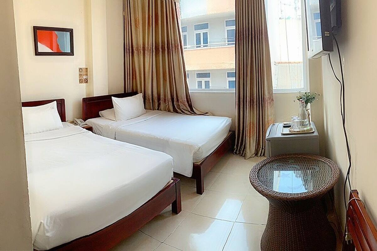 Quarto de hotel com duas camas, uma mesa pequena com um vaso e uma televisão na parede.