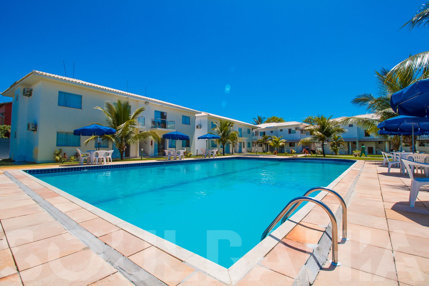 Uma piscina com um deck ladrilhado, cercada por palmeiras e guarda-sóis azuis.