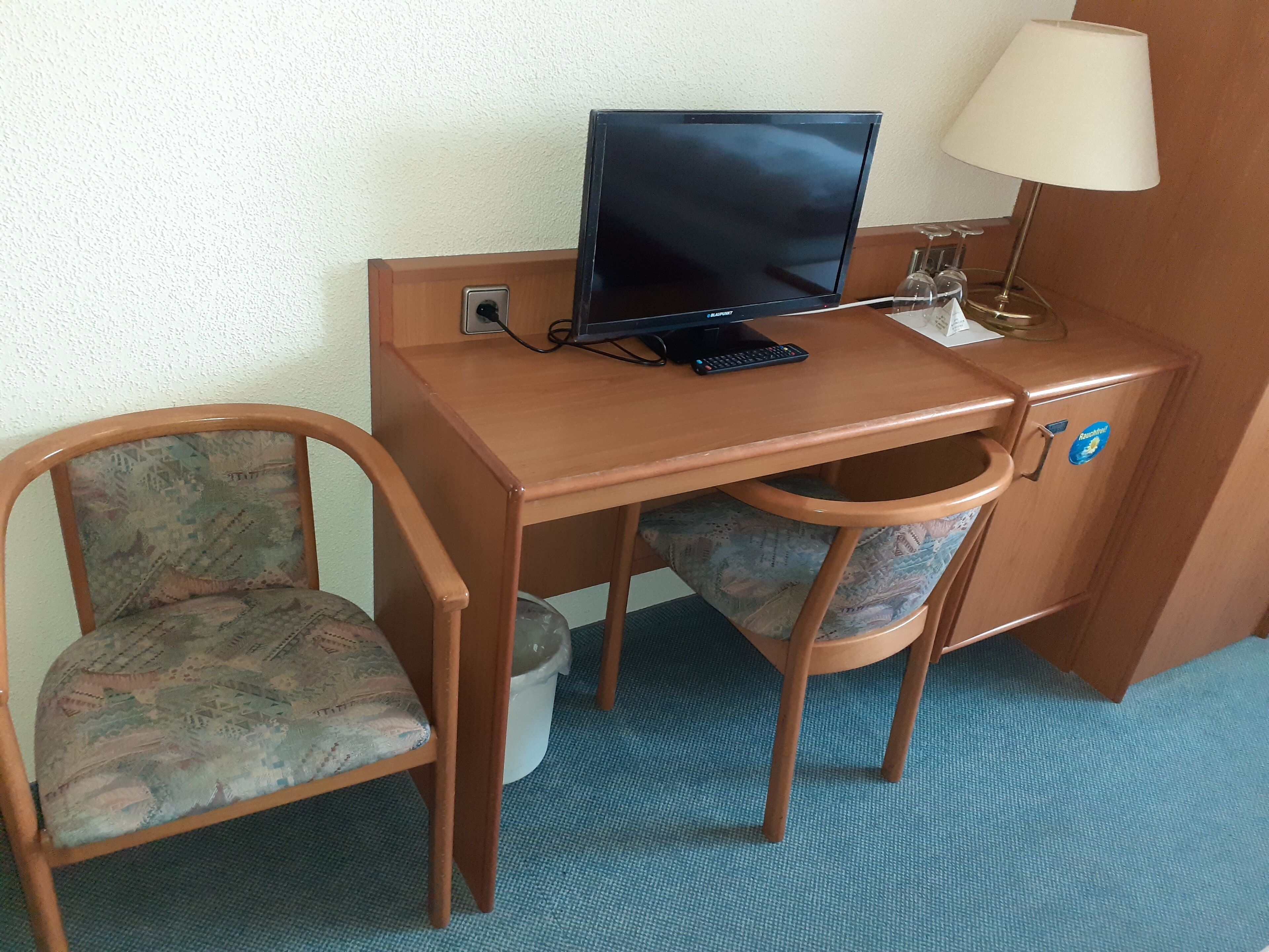 Uma mesa de madeira com um laptop, uma luminária e uma cadeira em um quarto com carpete azul.