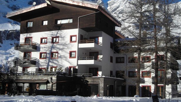 Hotel de vários andares com varandas, com uma paisagem de montanhas cobertas de neve ao fundo.