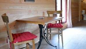 Interior de madeira com uma mesa e duas cadeiras, uma janela com cortinas e um candeeiro fixado na parede.