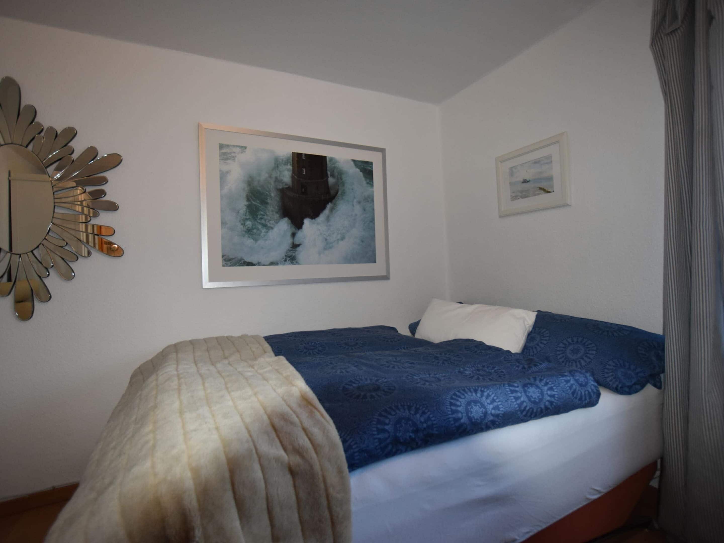 Quarto com cama, um quadro de farol e um espelho em formato de sol na parede.