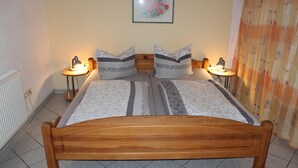 Cama bem arrumada com estrutura de madeira, duas mesas de cabeceira com abajures, um quadro floral na parede e uma cortina estampada.