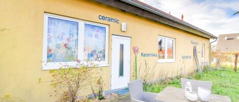 Um edifício amarelo com a palavra "ceramic" na fachada, uma pequena área externa com cadeiras de vime e uma mesa, e um jardim com plantas e flores.