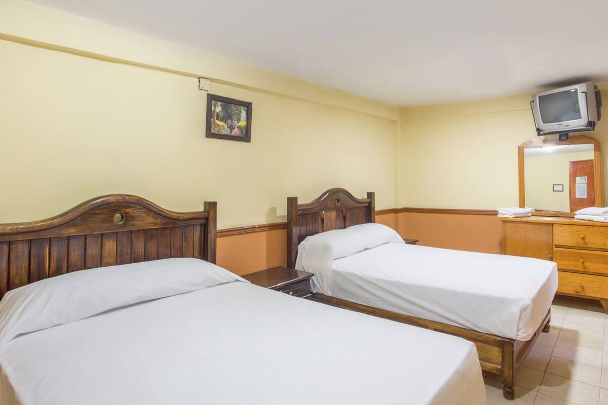 Quarto de hotel com duas camas de madeira, cômoda de madeira, televisão e quadro pendurado na parede.