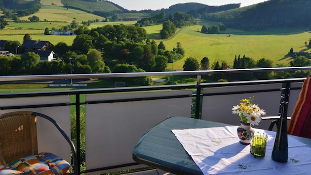 Uma varanda com cadeira de vime, uma pequena mesa com um vaso de flores, e vista para campos verdes e colinas.