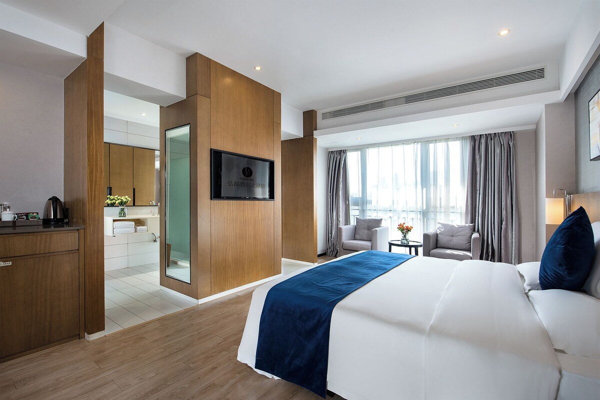 Quarto de hotel com cama, escrivaninha, cadeira, mesinha e janela com cortinas.