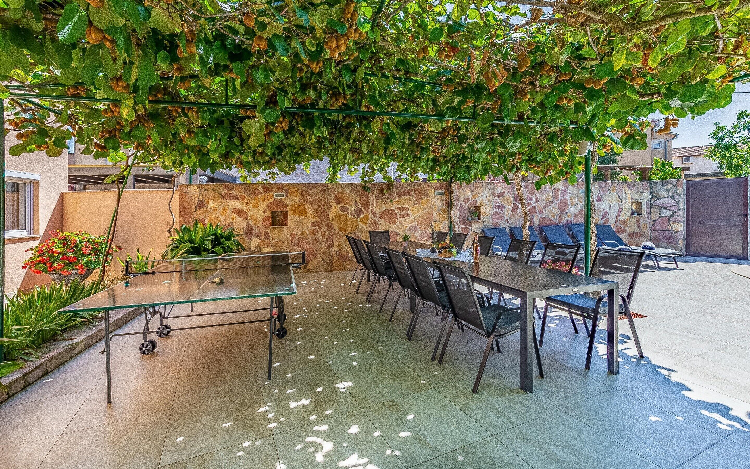 Um pátio externo com mesa e cadeiras de vidro, rodeado por paredes de pedra e vegetação.