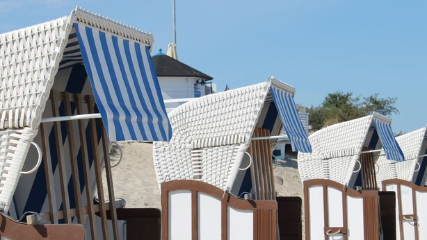 Uma fileira de cabanas de praia com toldos listrados, portas de madeira e uma praia arenosa ao fundo.