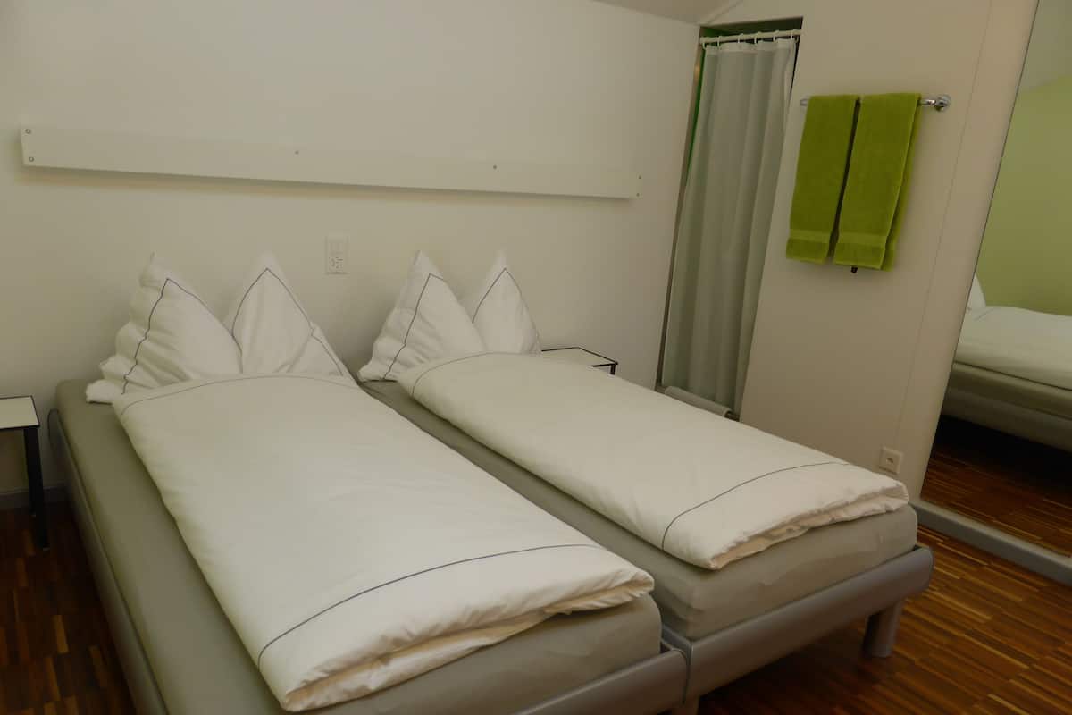 Quarto duplo com duas camas de solteiro, piso de madeira, toalha verde e janela com cortina.
