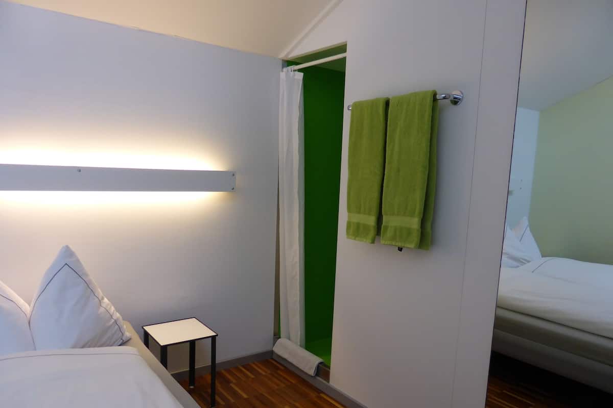 Quarto de hotel com uma cama, uma mesinha, uma toalha verde e uma cortina branca.
