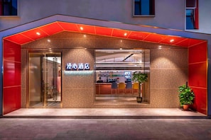 Entrada de hotel com design moderno, apresentando esquema de cores em vermelho e bege, porta de vidro e placa com o nome do hotel.