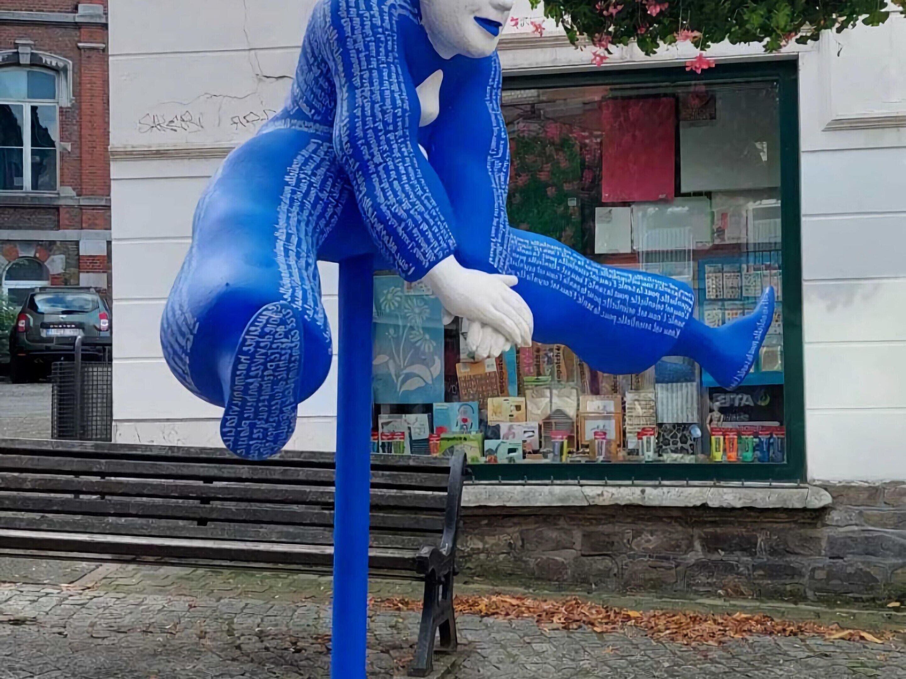 Escultura azul de uma figura com texto no corpo, posicionada em frente a um edifício com uma janela exibindo diversos objetos.