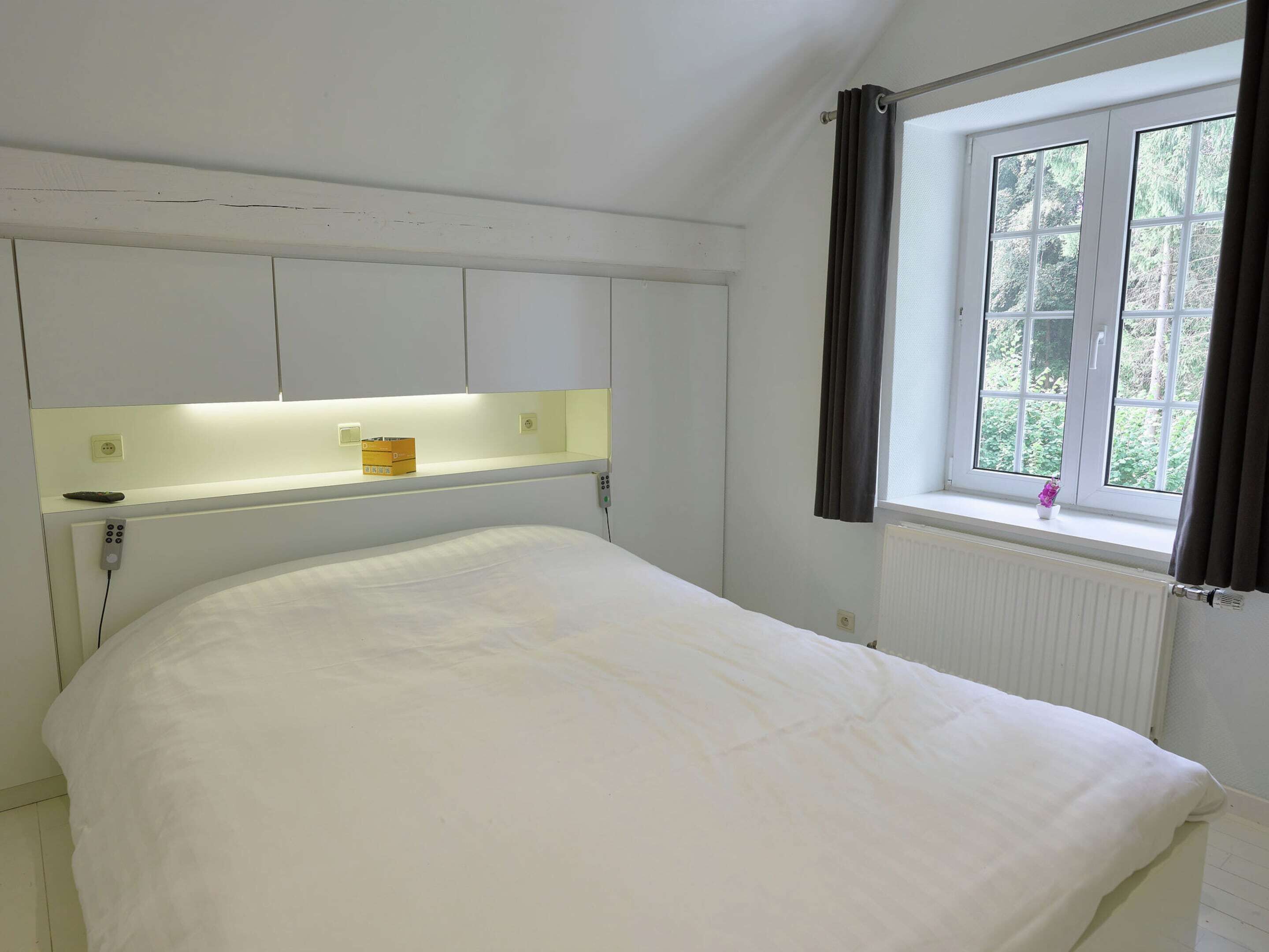 Um quarto com uma cama, uma janela com cortinas, uma prateleira com uma caixa e um painel de controle.
