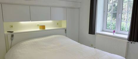 Um quarto com uma cama, uma janela com cortinas, uma prateleira com uma caixa e um painel de controle.