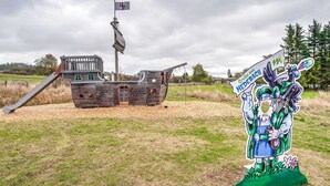 Uma estrutura de recreação em forma de barco de madeira, com um escorregador e uma bandeira, situada num campo relvado com um painel de personagem de desenho animado nas proximidades.