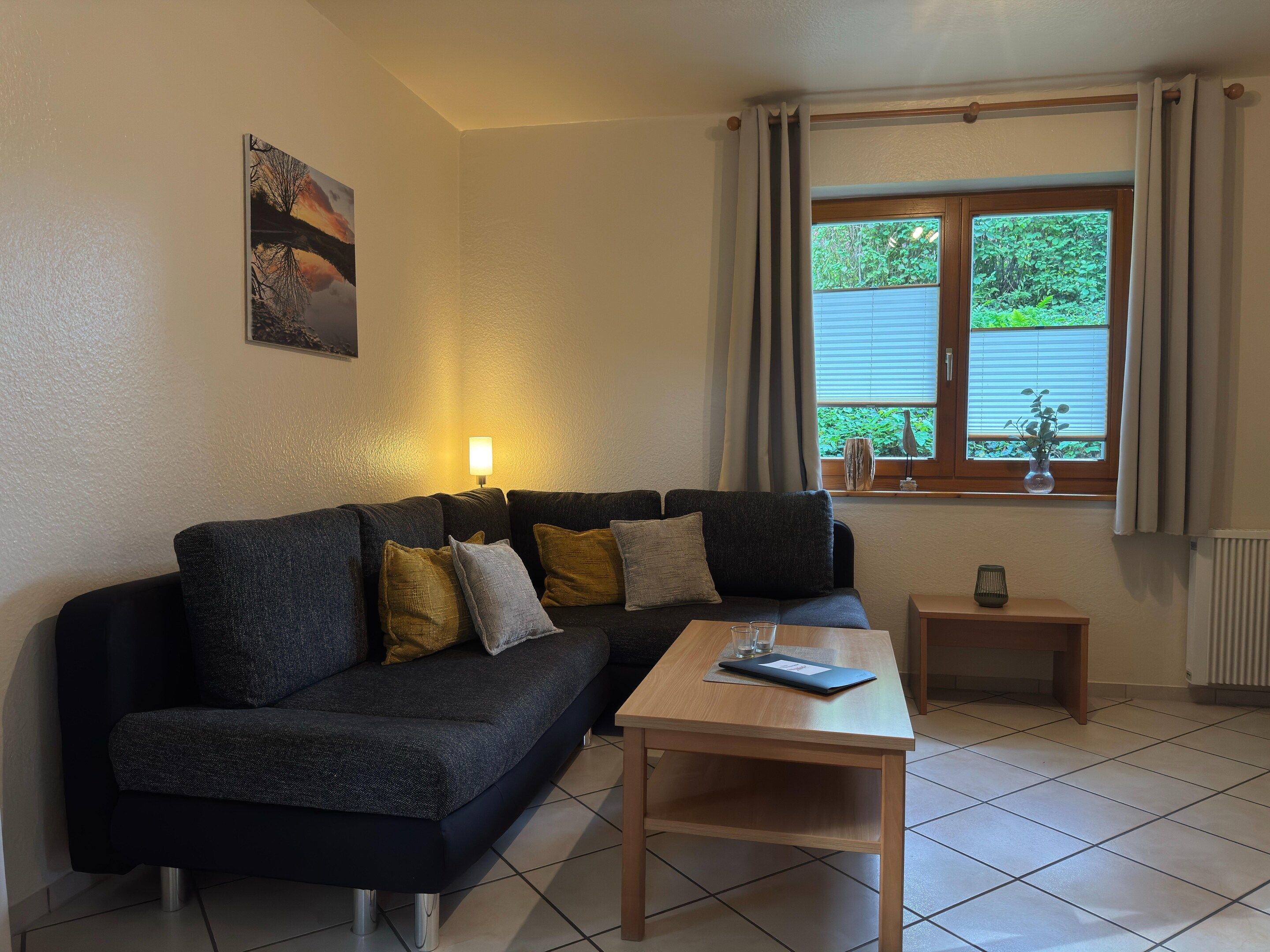 Sala de estar com sofá escuro, mesa de centro de madeira, luminária e janela com cortinas.