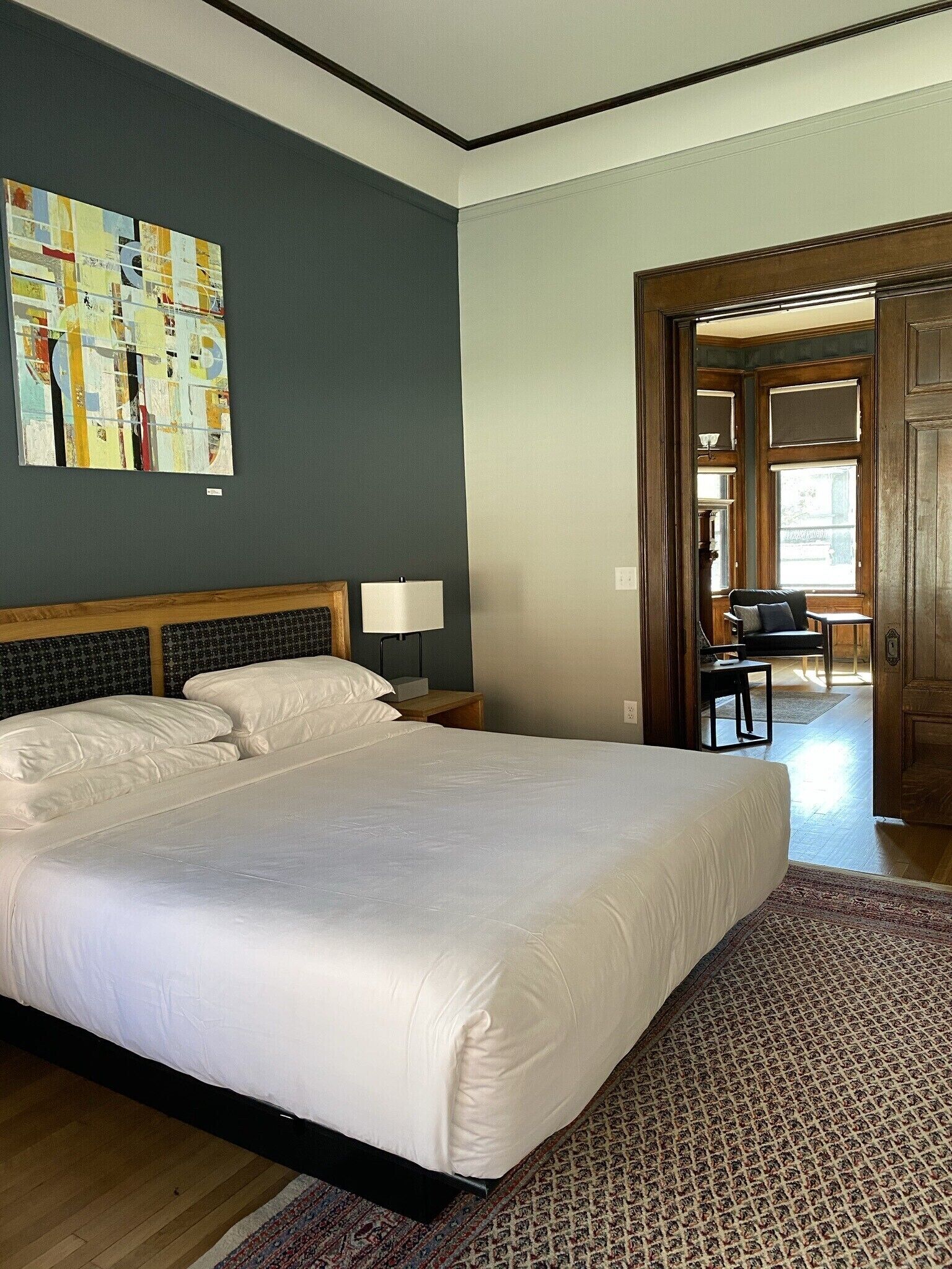 Quarto de hotel com uma cama grande, mesa de cabeceira com abajur, quadro na parede e vista para o exterior através de uma porta.