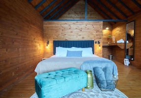 Quarto de cabana de madeira com uma cama, um banquinho estofado azul e uma pequena mesa de cabeceira.
