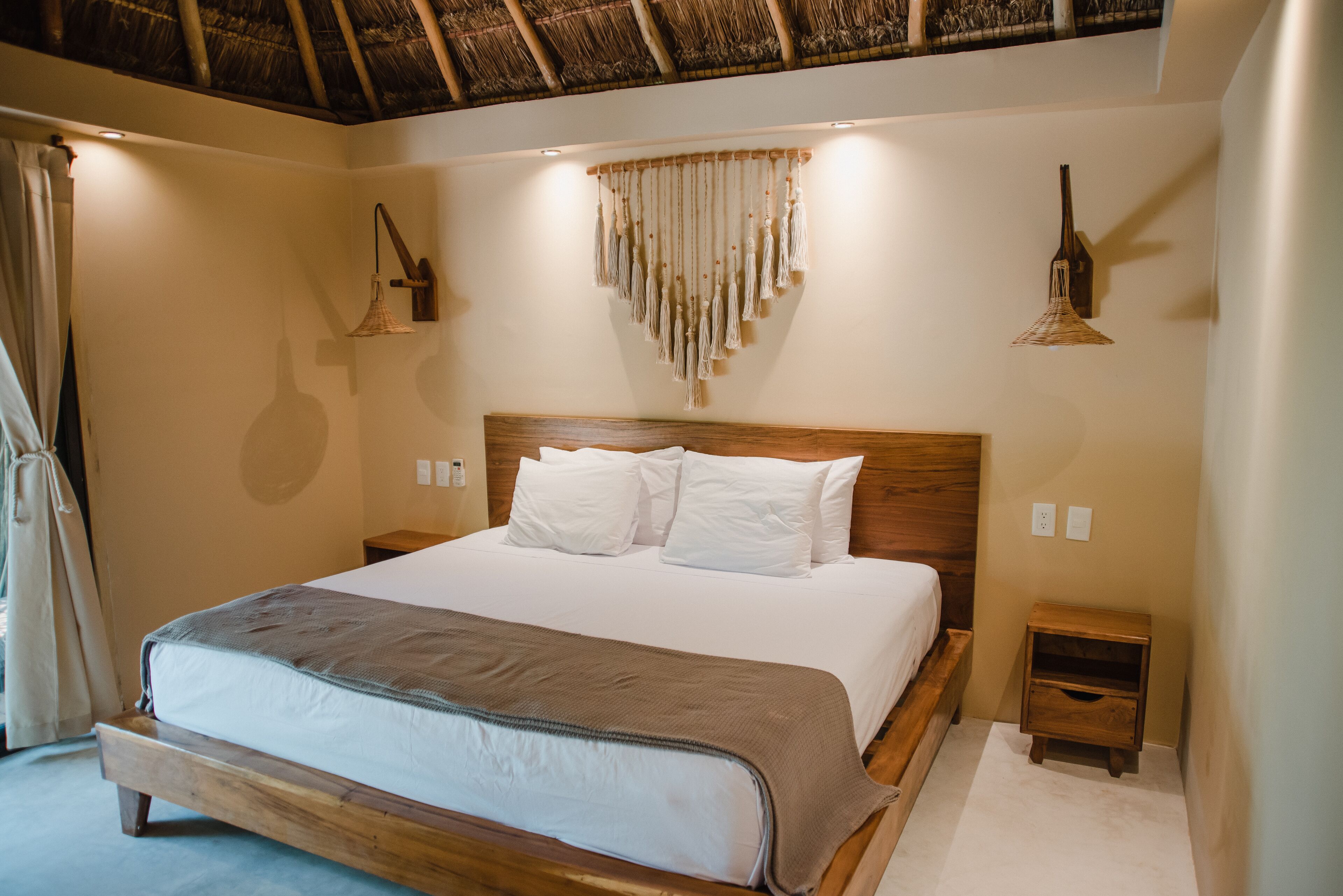 Quarto com cama de madeira, roupa de cama branca, decoração de macramé pendente e uma pequena mesa de cabeceira de madeira.