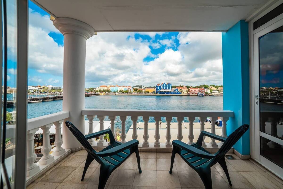 Uma varanda com duas cadeiras, vista para uma orla com edifícios coloridos e céu claro.