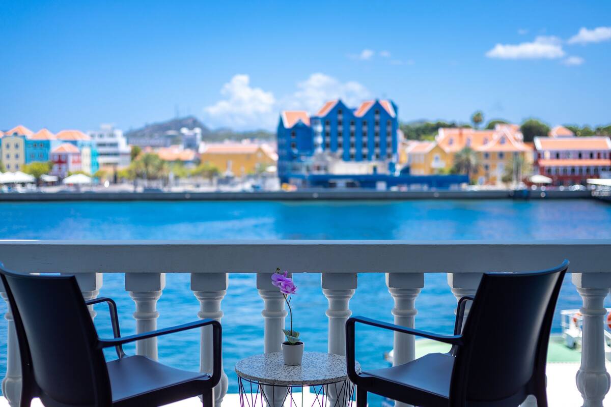 Varanda com duas cadeiras e uma pequena mesa, com vista para uma orla com edifícios coloridos.