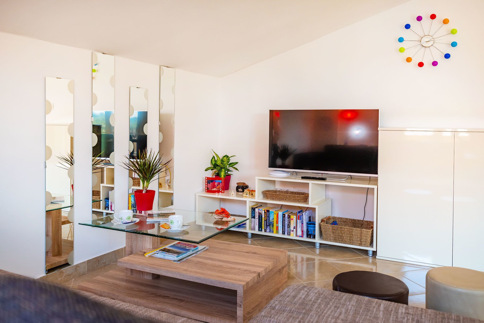Uma sala de estar moderna com uma mesa de centro de vidro, uma TV de tela plana, uma estante e plantas decorativas.