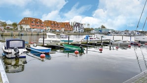 Uma marina com vários barcos atracados, um edifício com telhados vermelhos e água calma refletindo o céu.
