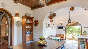 Uma sala de jantar com uma mesa de madeira, cadeiras azuis e uma tigela de frutas. Há um armário de madeira com prateleiras, uma televisão e um ventilador de teto.