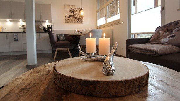 Uma sala de estar moderna com uma mesa de madeira redonda, duas velas acesas e um vaso de prata decorativo.