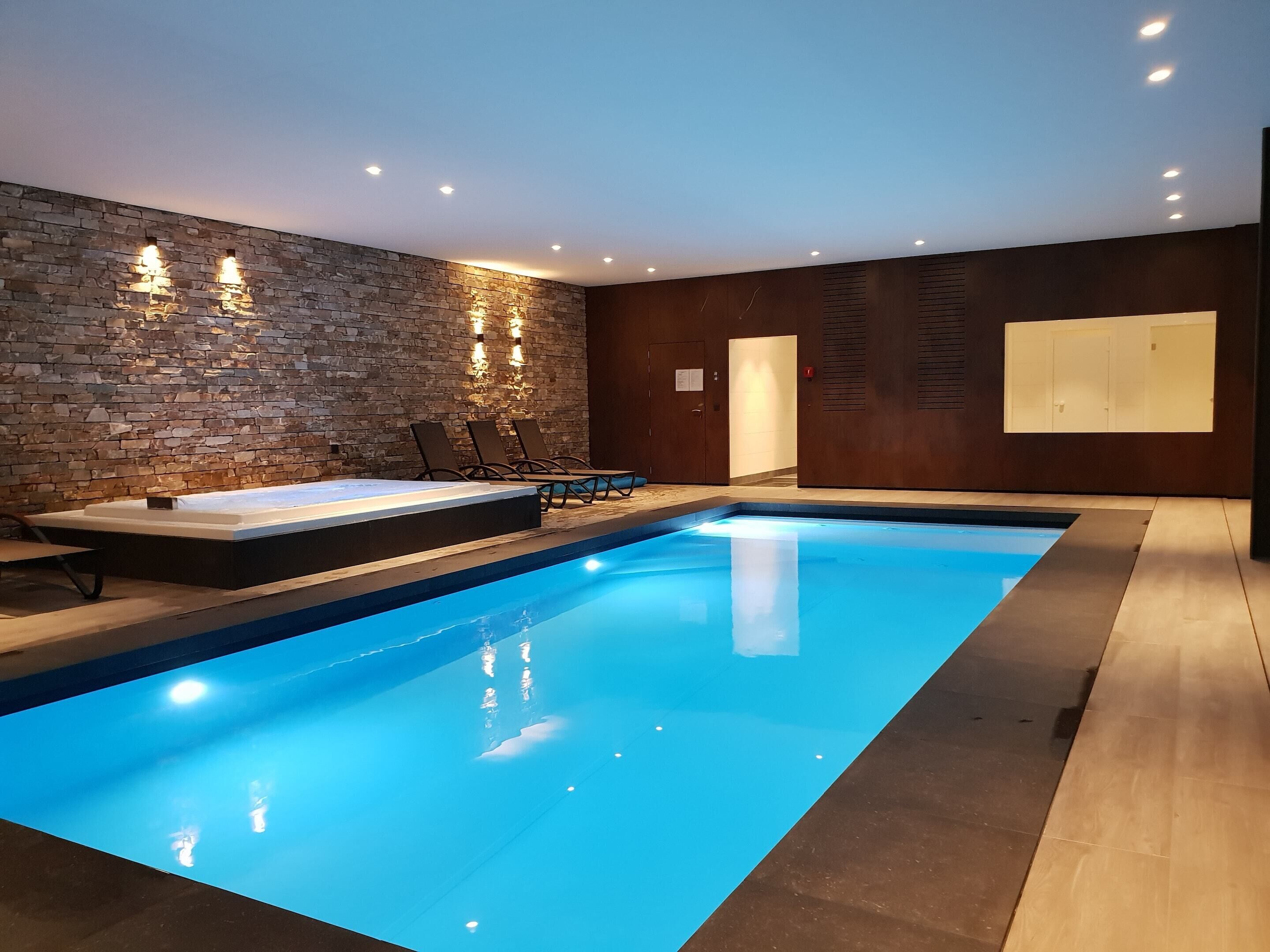 Uma piscina com paredes de pedra e madeira, duas espreguiçadeiras e uma mesa para massagem.