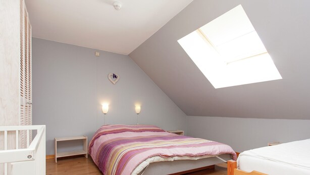 Um quarto com teto inclinado, um claraboia, uma cama com colcha listrada e colorida, uma mesinha de cabeceira e um banco de madeira.