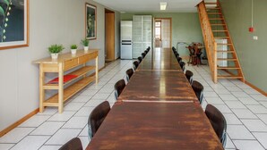 Uma longa mesa de madeira com cadeiras, um banco de madeira, um refrigerador e uma escada.