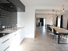 Uma cozinha moderna com painel de azulejos pretos, armários brancos e uma mesa de jantar comprida com cadeiras.