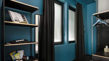 Quarto de hotel com parede azul, janela com cortinas, cama, prateleira com secador de cabelo e pequena mesa com artigos de higiene pessoal.