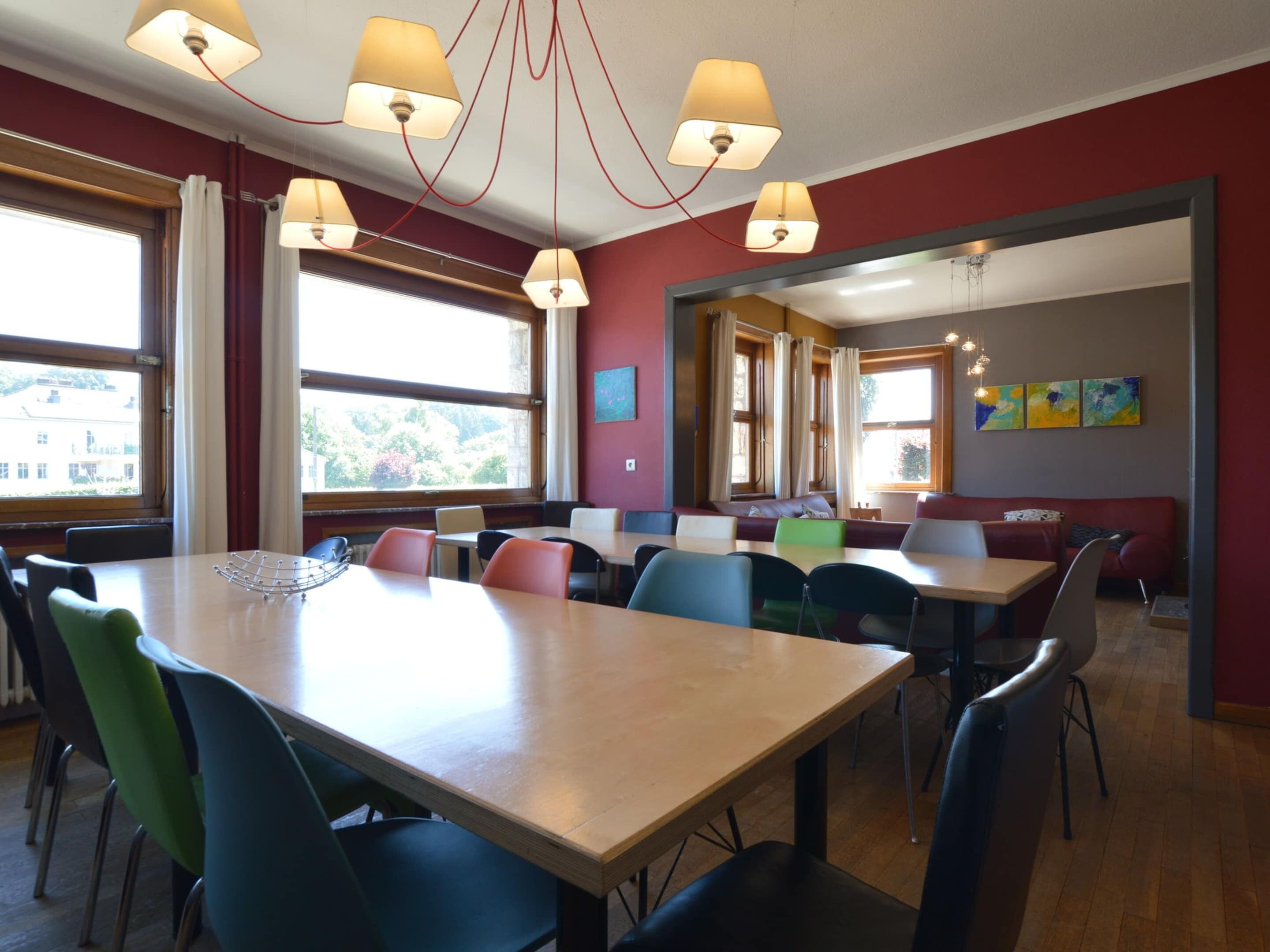 Uma sala de jantar com uma mesa grande e várias cadeiras, janelas amplas com cortinas e uma parede vermelha com arte abstrata.