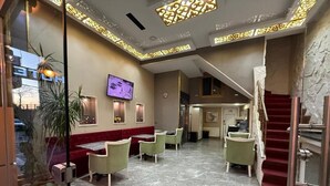 Um saguão de hotel com piso de mármore, cadeiras verdes, um sofá vermelho, uma escadaria e uma televisão na parede.