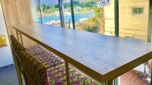 Uma mesa de madeira e um banco de palha em um quarto com grandes janelas com vista para um corpo de água e o letreiro de um hotel.