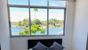 Quarto com cama, janela com vista para um lago e vegetação, e uma porta.