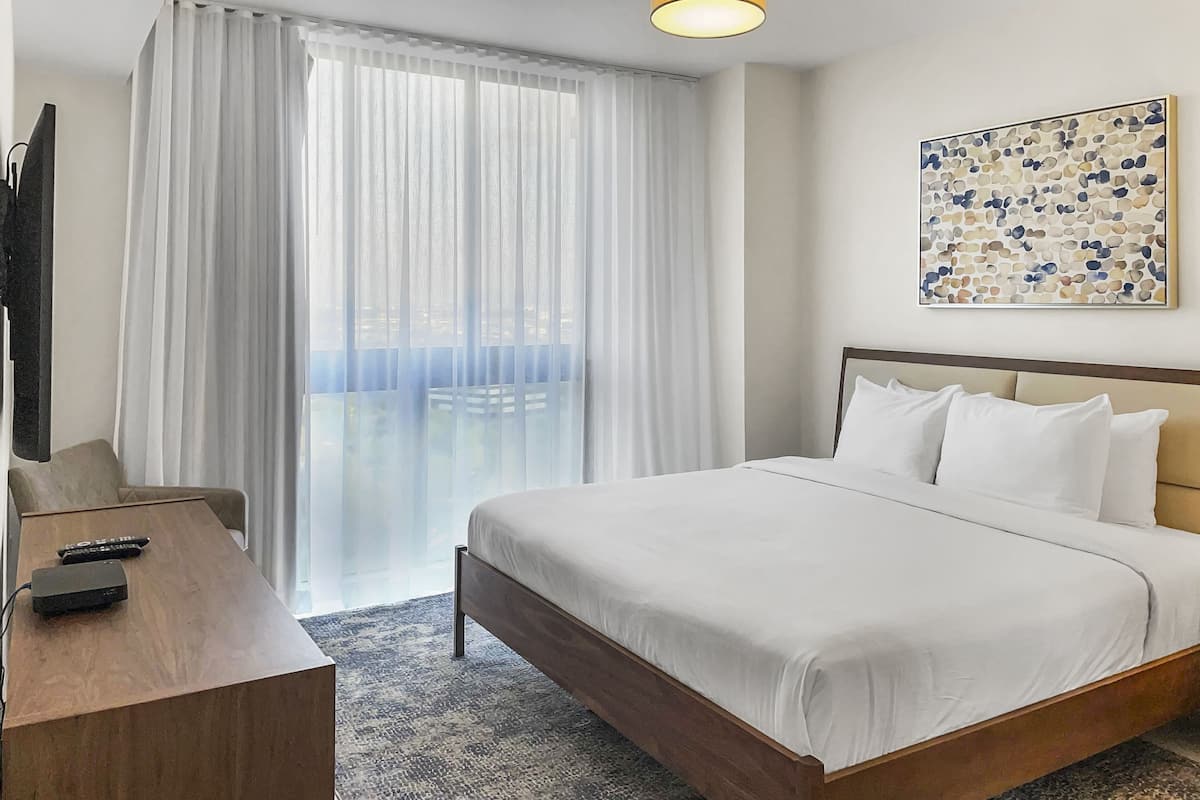Quarto de hotel com uma cama grande, uma escrivaninha com televisão e controles remotos, um quadro na parede e vista através de cortinas translúcidas.