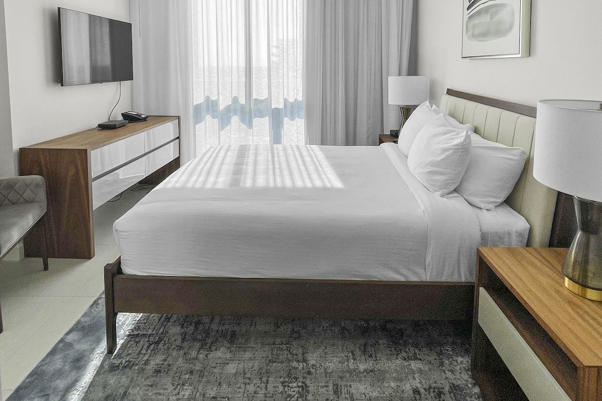 Quarto de hotel moderno com uma cama grande, televisão de tela plana, mesa de cabeceira de madeira, escrivaninha e uma janela ampla com cortinas finas.