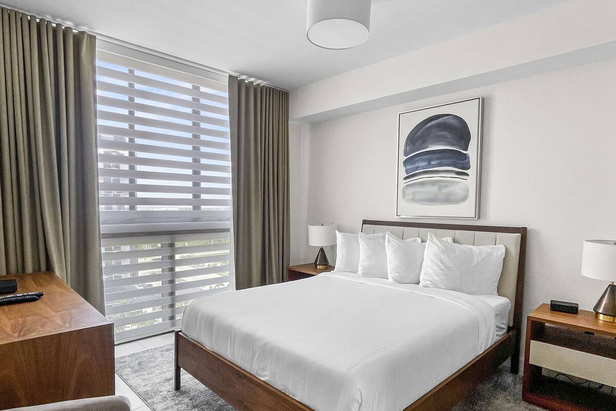 Quarto de hotel com uma cama grande, uma escrivaninha, uma cadeira, mesa de cabeceira e uma janela com persianas.
