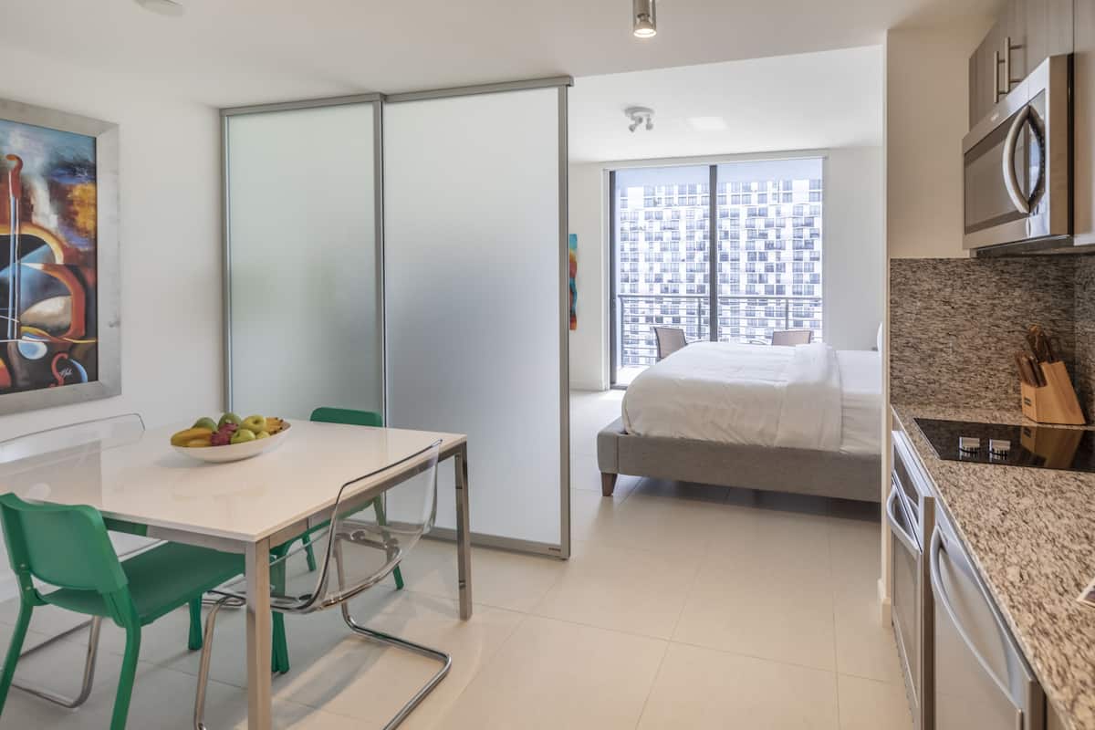 Quarto de hotel moderno com cama, área de jantar com mesa e cadeiras, cozinha com micro-ondas e uma janela grande com vista para a cidade.